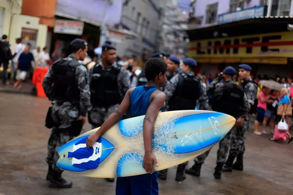 Knabo kun ĉena tabulo rigardas la policon, Rosigna, Rio de Janeiro, Brazilo. Unu el la plej malfacilaj taskoj de Brazilo antaŭ la 2014-datita FIFA-Monda Pokalo estas senvestigado de gravaj krimaj areoj.